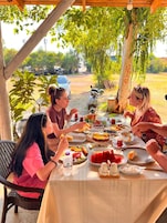 Um grupo de pessoas jantando ao ar livre em uma mesa com diversos pratos, sob uma pérgola de madeira com árvores ao fundo.