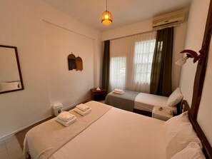Quarto de hotel com uma cama grande, uma cama menor, uma mesinha de cabeceira com abajur, um espelho e uma janela com cortinas.
