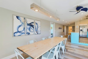 Uma sala de jantar com mesa de madeira e cadeiras brancas, uma cozinha ao fundo e arte abstrata na parede.