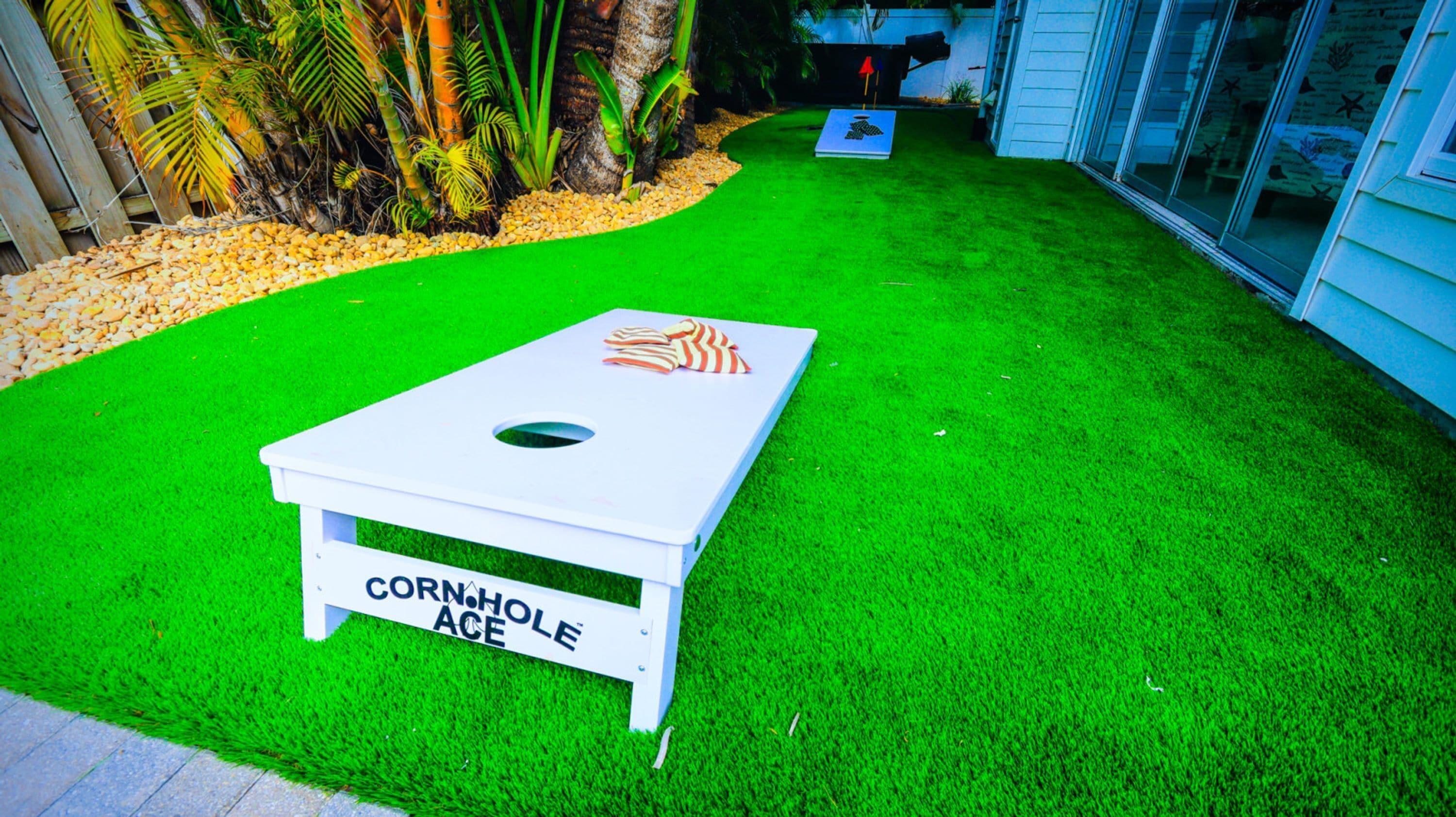Jogo de cornhole montado em grama sintética, com uma mesa branca e uma bandeira vermelha.