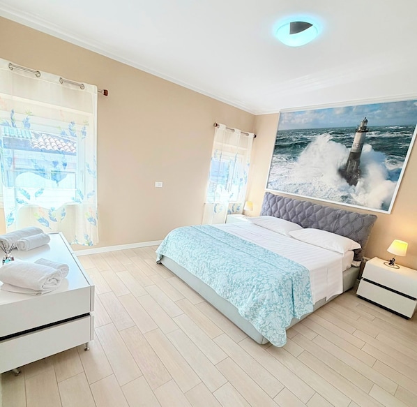 Quarto com cama, mesa de cabeceira, cortinas na janela, um quadro na parede e luz no teto.
