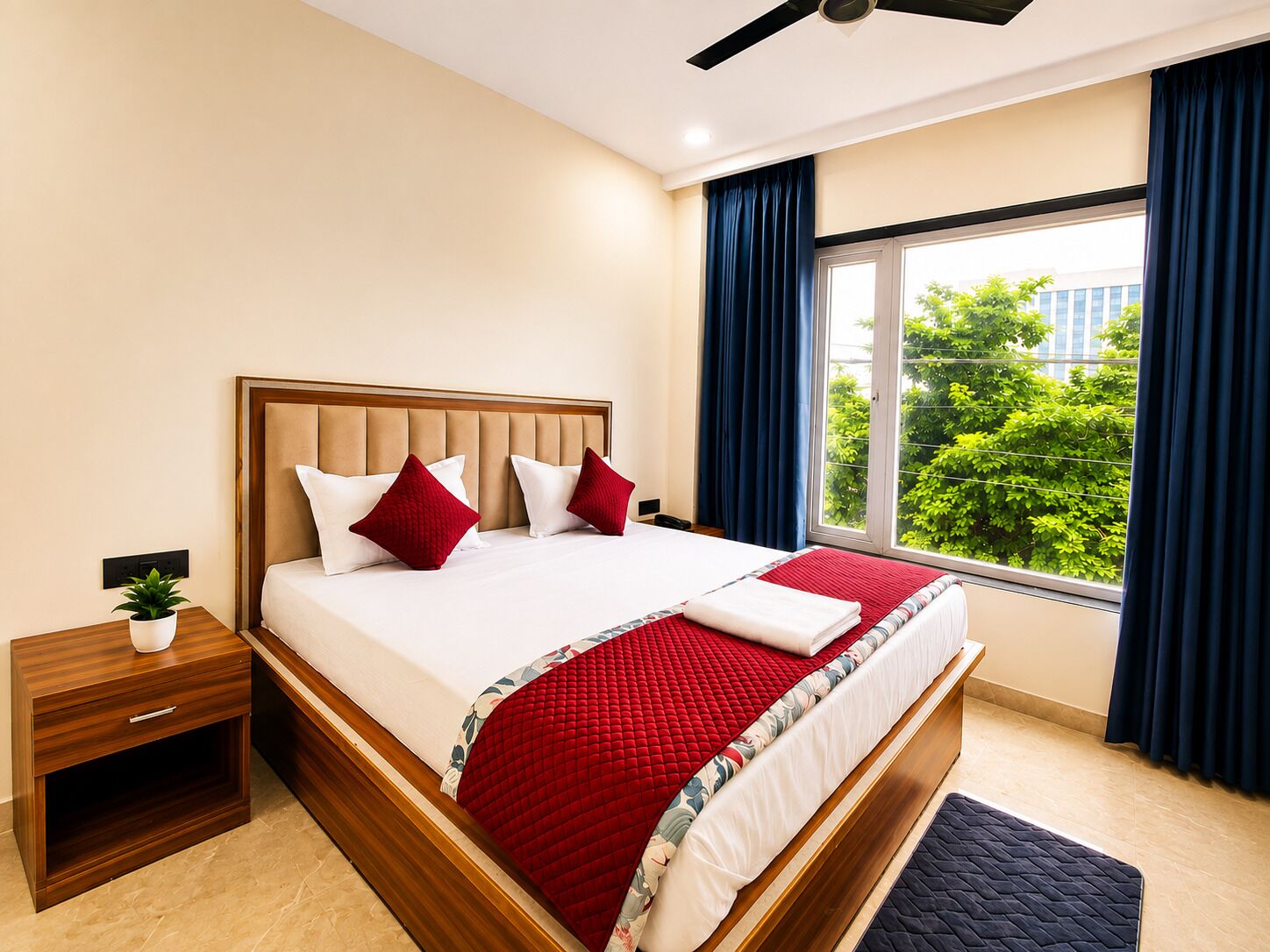 Quarto de hotel com cama, mesa de cabeceira, planta em vaso e janela com cortinas.