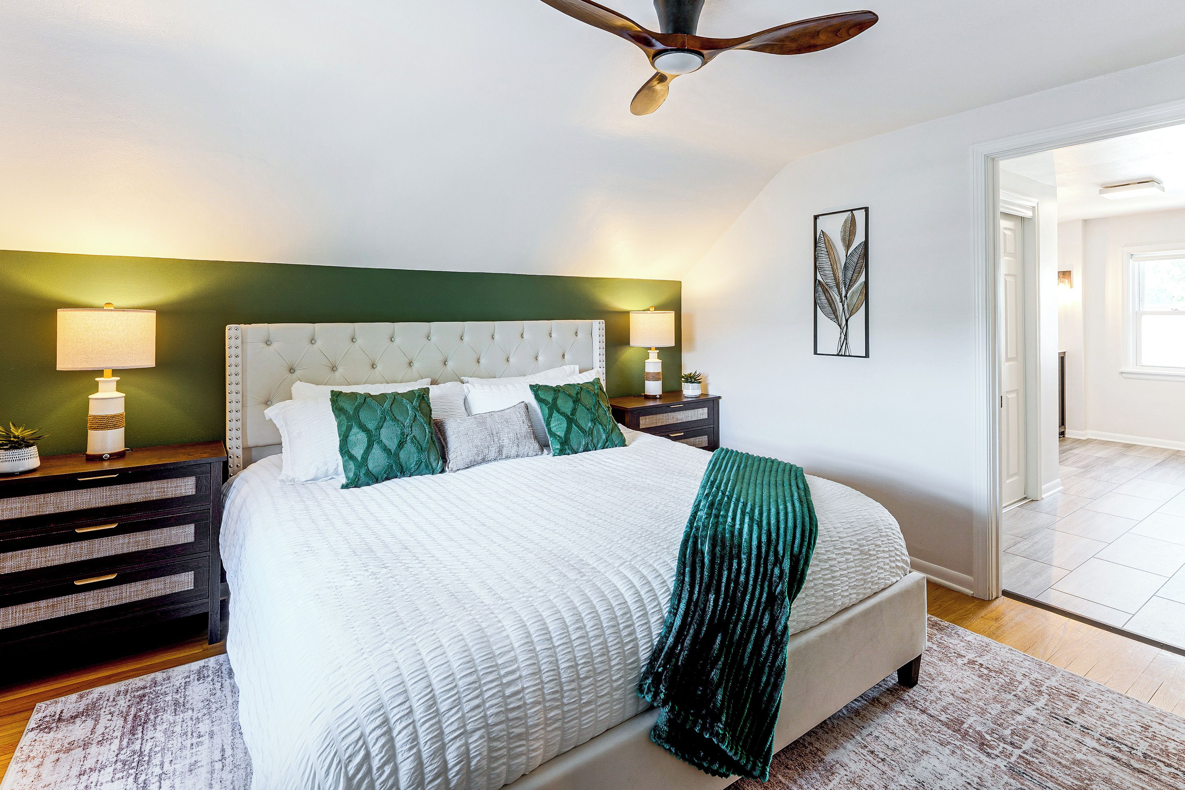 Quarto com cabeceira estofada, cama com roupa de cama branca e almofadas verdes, mesa de cabeceira com abajur e ventilador de teto.