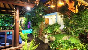 Um jardim tropical com piscina, deck de madeira e um edifício com varanda.