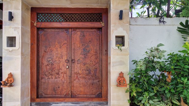 Uma porta de madeira grande com entalhes elaborados, ladeada por duas pequenas estátuas e um vaso de planta.