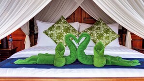 Uma cama com lençóis brancos, duas almofadas verdes com padrão de folha e uma toalha verde disposta em formato de coração.