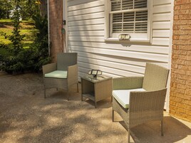 Uma área de estar externa com móveis de vime e uma mesa pequena, ao lado de uma casa com janela e parede de tijolos.