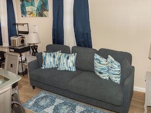 Um sofá cinzento com almofadas estampadas, uma mesinha com uma lâmpada, uma cafeteira e um quadro abstrato pendurado na parede.