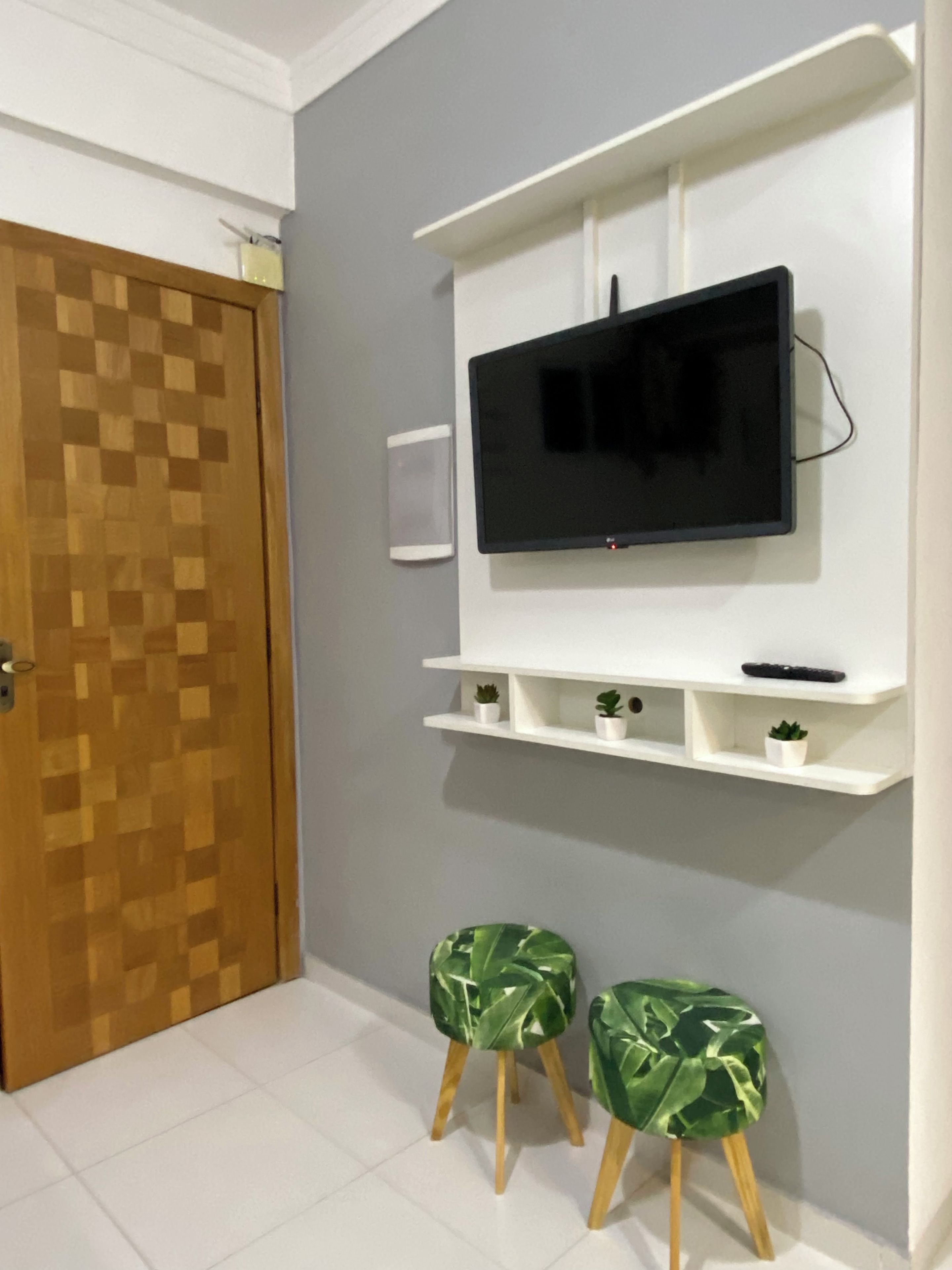 Quarto com televisão presa a uma parede branca, porta de madeira com padrão pixelado e dois banquinhos verdes com pés de madeira.