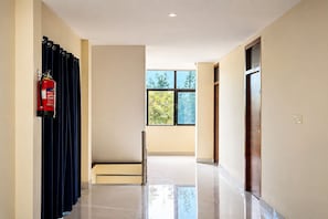 Um corredor limpo e moderno, com um extintor preso na parede, uma janela com vista para o verde, e uma porta que dá para outro cômodo.