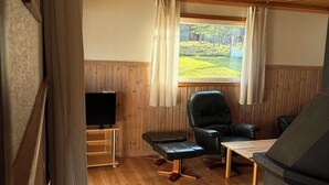 Um quarto aconchegante com piso de madeira, televisão, poltrona de couro, mesa pequena e janela com cortinas.