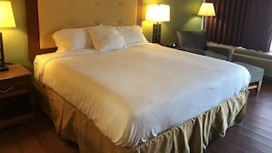 Uma cama grande com cabeceira de madeira, duas luminárias de cabeceira, uma mesinha e uma cadeira estampada.