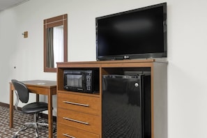 Quarto de hotel com escrivaninha de madeira, uma cadeira preta, micro-ondas, geladeira e televisão sobre um suporte.