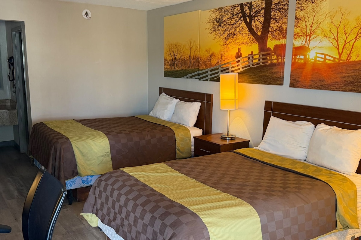 Quarto de hotel com duas camas, uma escrivaninha, uma cadeira e uma luminária fixada na parede.