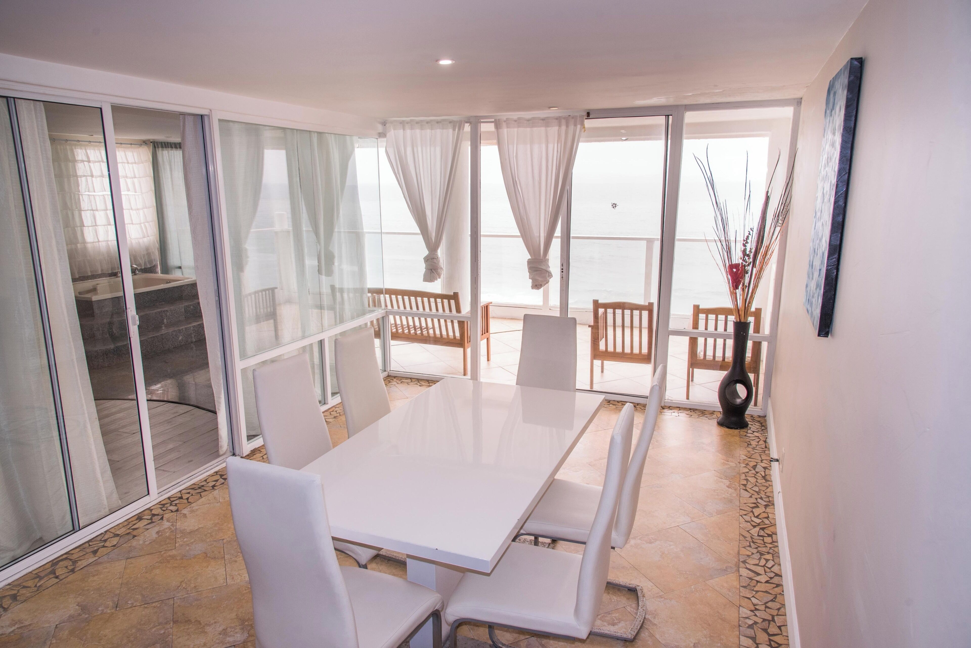 Uma área de refeições com mesa e cadeiras brancas, vista para o mar através de grandes janelas e uma varanda com cadeiras de madeira.