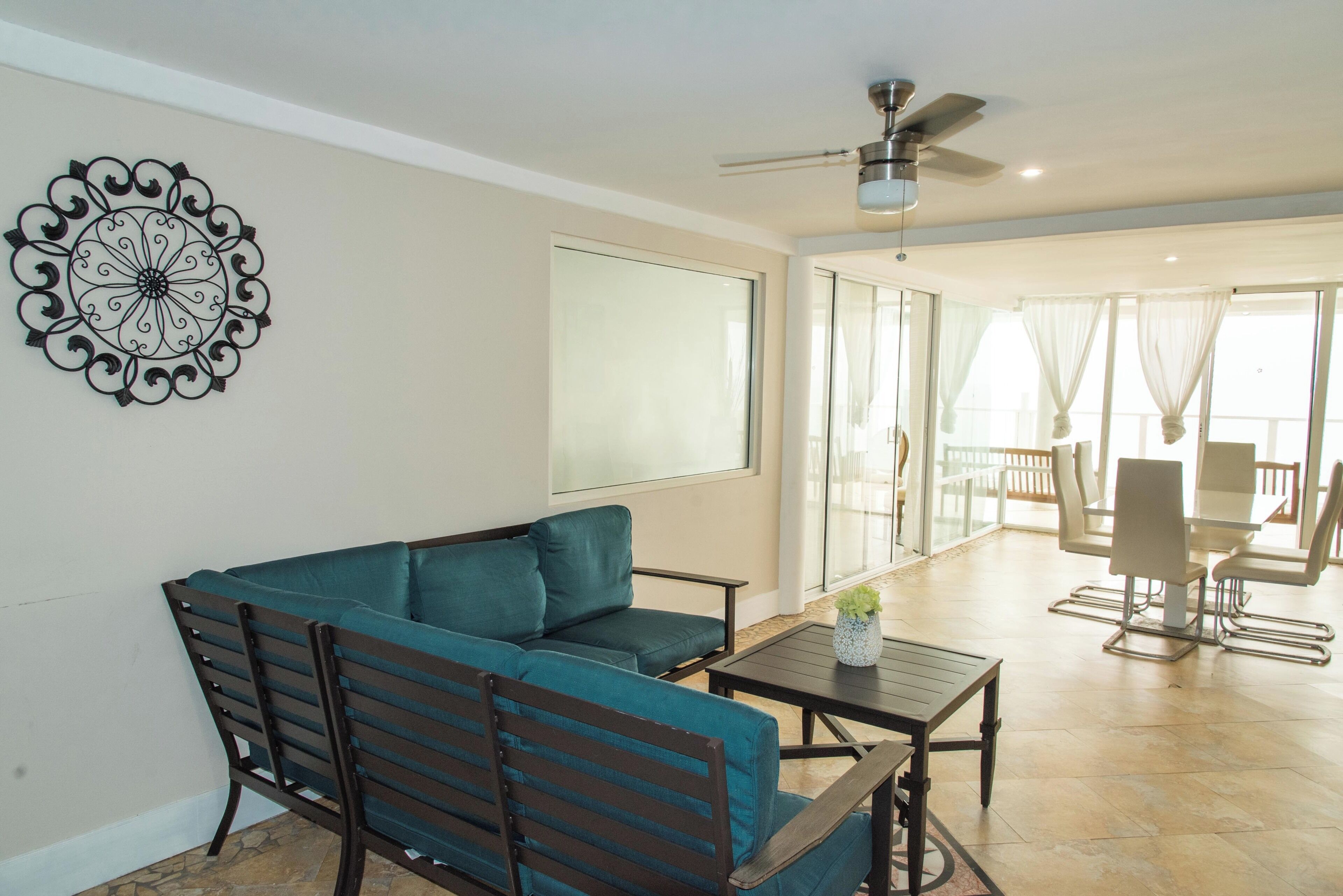 Sala de estar com sofá, mesa de centro e um objeto decorativo na parede. Há um ventilador de teto e vista para uma varanda com mobiliário exterior.