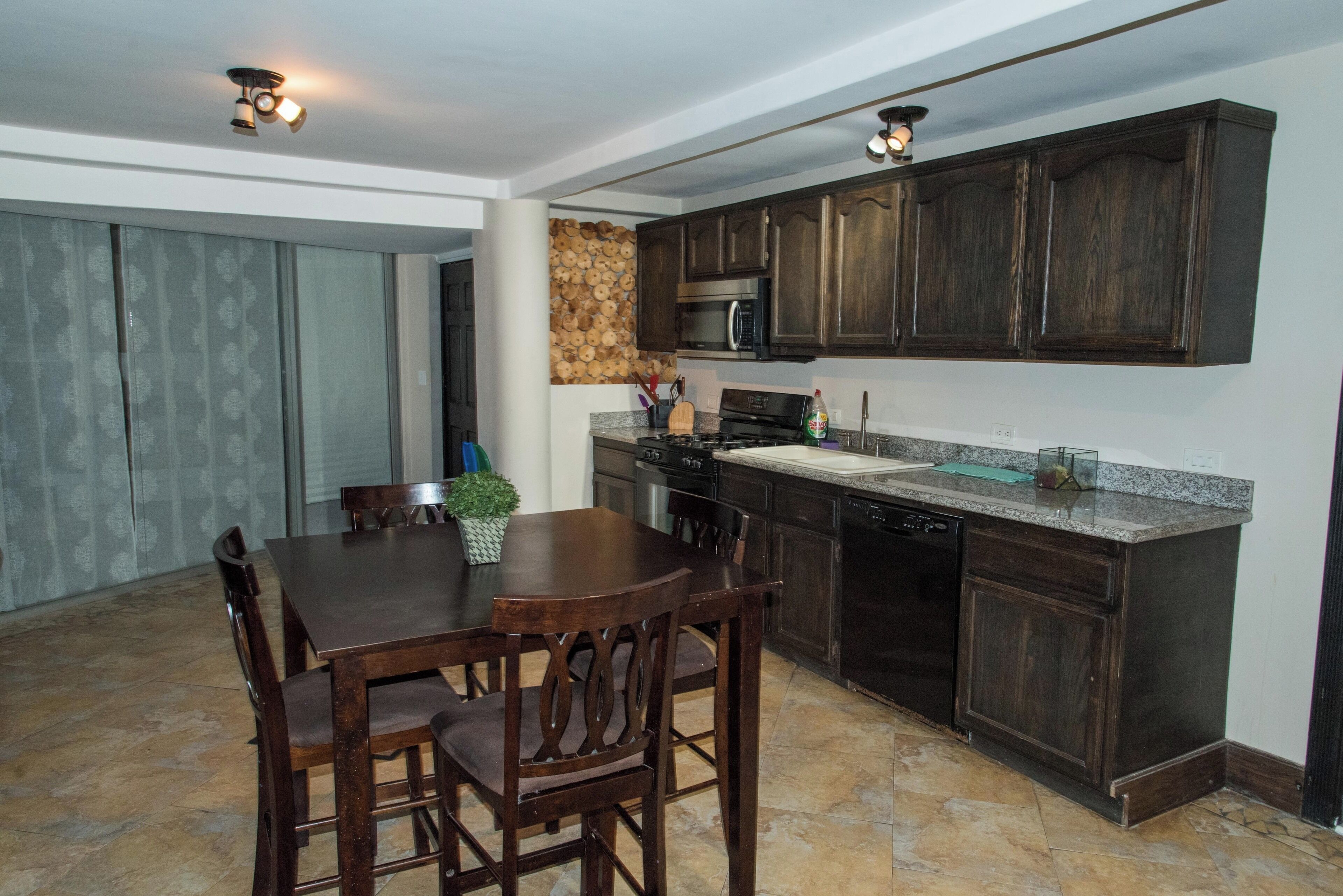 Cozinha com armários de madeira escura, bancada de granito, micro-ondas embutido e lava-louças. Há uma mesa de jantar com cadeiras e um vaso de planta.