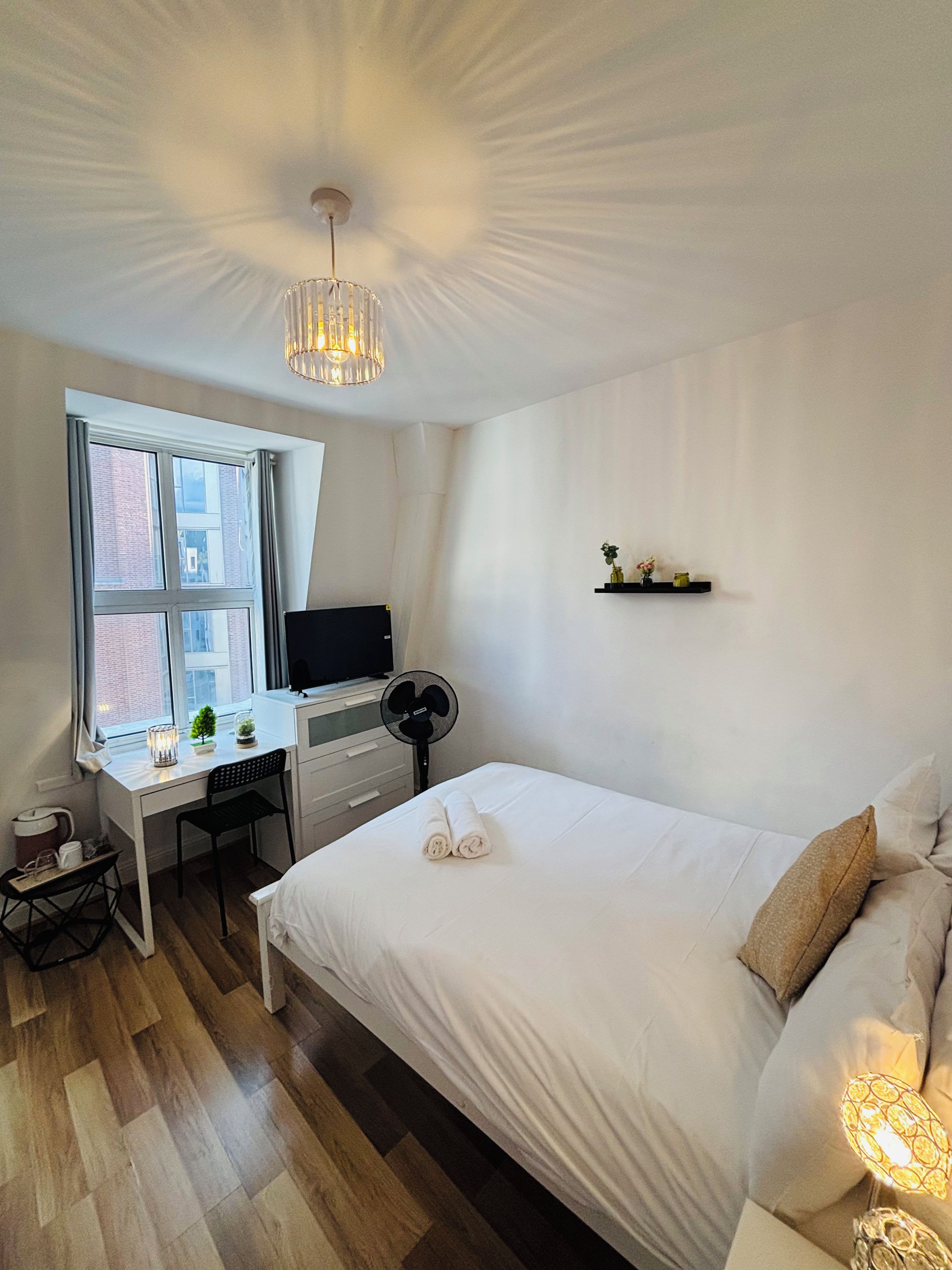 Quarto de hotel com uma cama, uma mesa de trabalho com computador, uma cadeira, uma janela e um ventilador de teto.