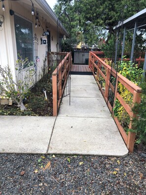Um caminho de concreto leva a um deck de madeira com guarda-corpo de metal, cercado por vegetação e um edifício com janela e porta.