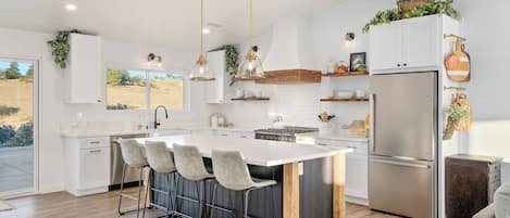 Uma cozinha moderna com uma ilha central, banquetas, eletrodomésticos de aço inoxidável e vista para o exterior.
