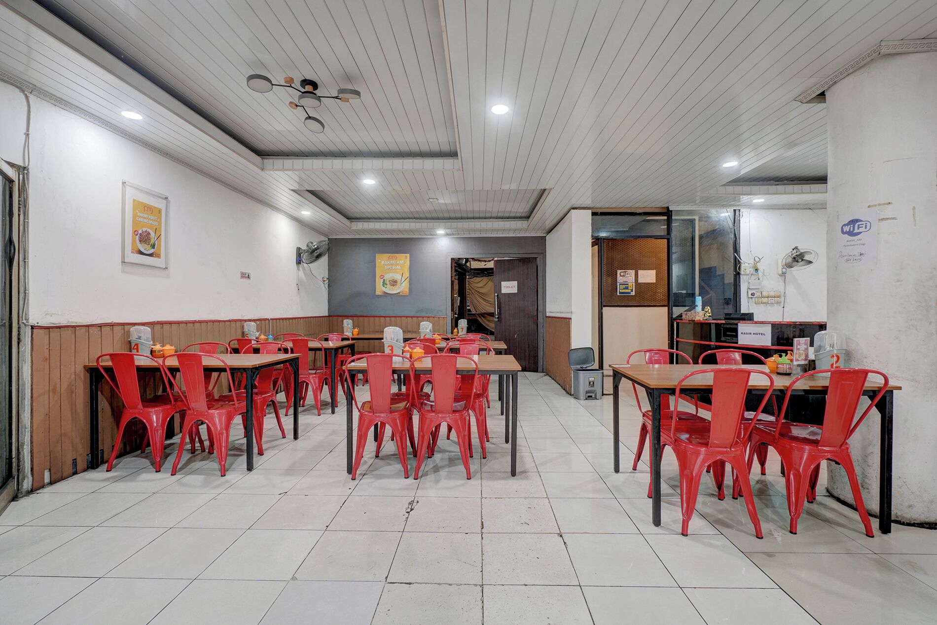 Uma área de refeições de hotel com cadeiras e mesas vermelhas, um ventilador de teto e uma placa com a inscrição 'KASIR HOTEL'.