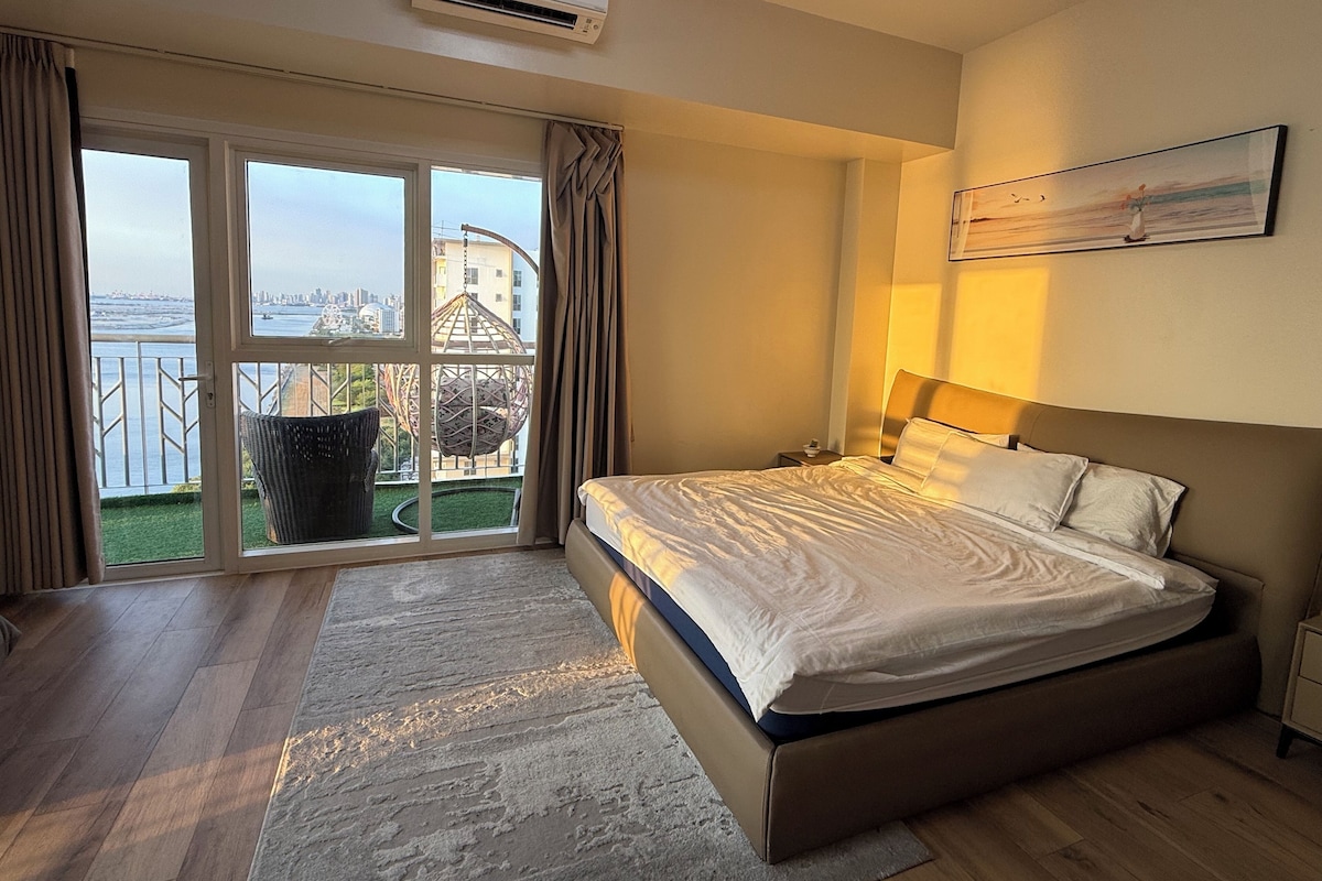Quarto com cama grande, vista da cidade através de uma porta de vidro e uma cadeira suspensa perto da porta.