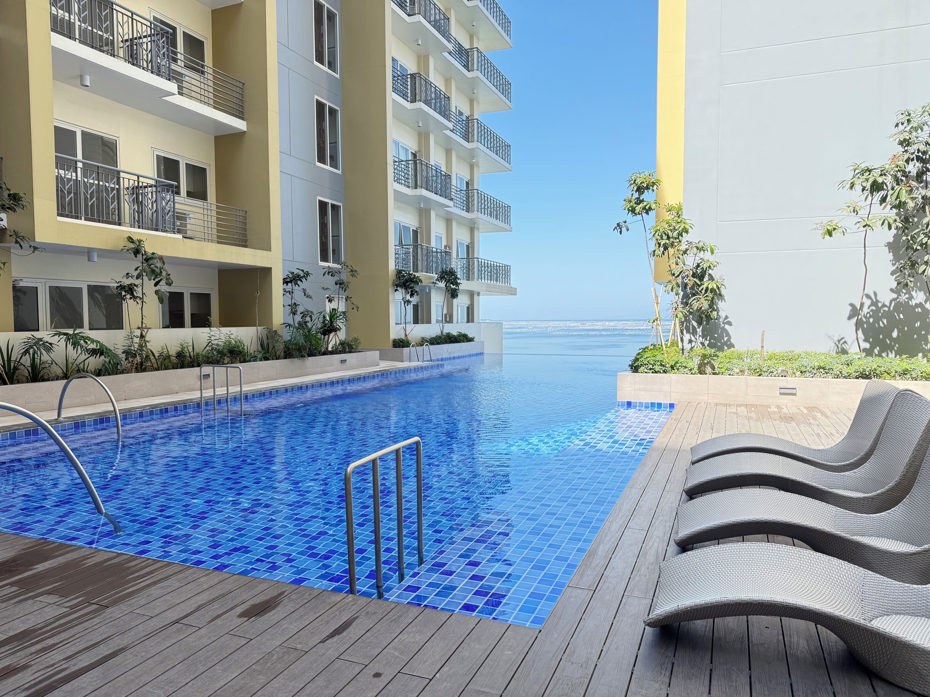 Piscina com deck de madeira e espreguiçadeiras, cercada por edifícios altos com sacadas e plantas.