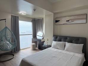 Quarto de hotel moderno com uma cama grande, uma cadeira suspensa, uma mesa de cabeceira com luminária, um espelho e vista para o mar pela janela.