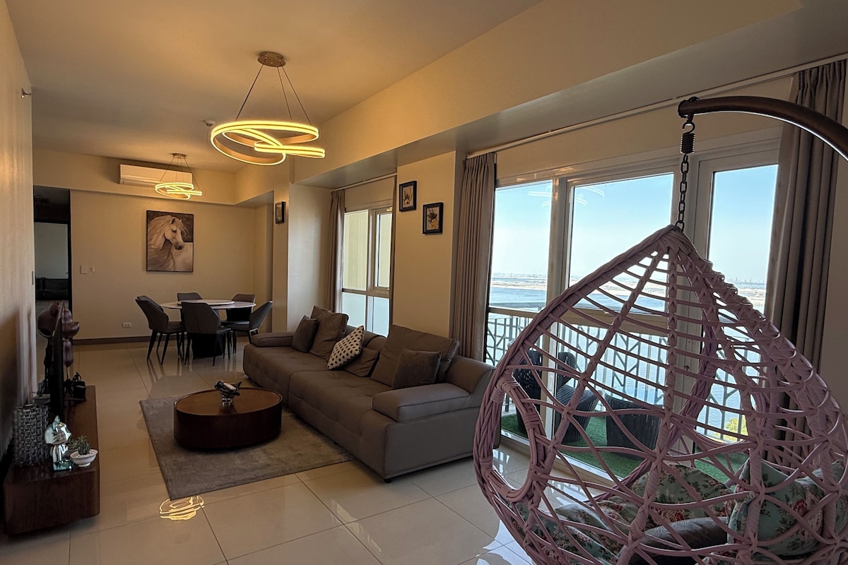 Uma sala de estar moderna com sofá, mesa de centro e cadeira suspensa, área de refeições com cadeiras e janelas amplas com vista.