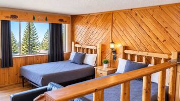 Quarto no estilo cabana com duas camas, cabeceira de madeira, mesa de cabeceira com abajur e vista para as árvores pela janela.
