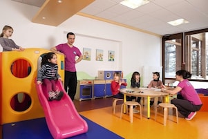 Espaço infantil com escorrega, mesa com brinquedos e janela com vista para árvores.