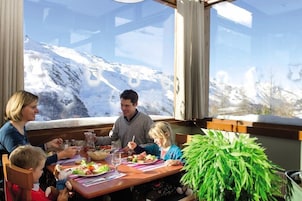 Uma família jantando à mesa com vista para uma paisagem montanhosa nevada através de grandes janelas.