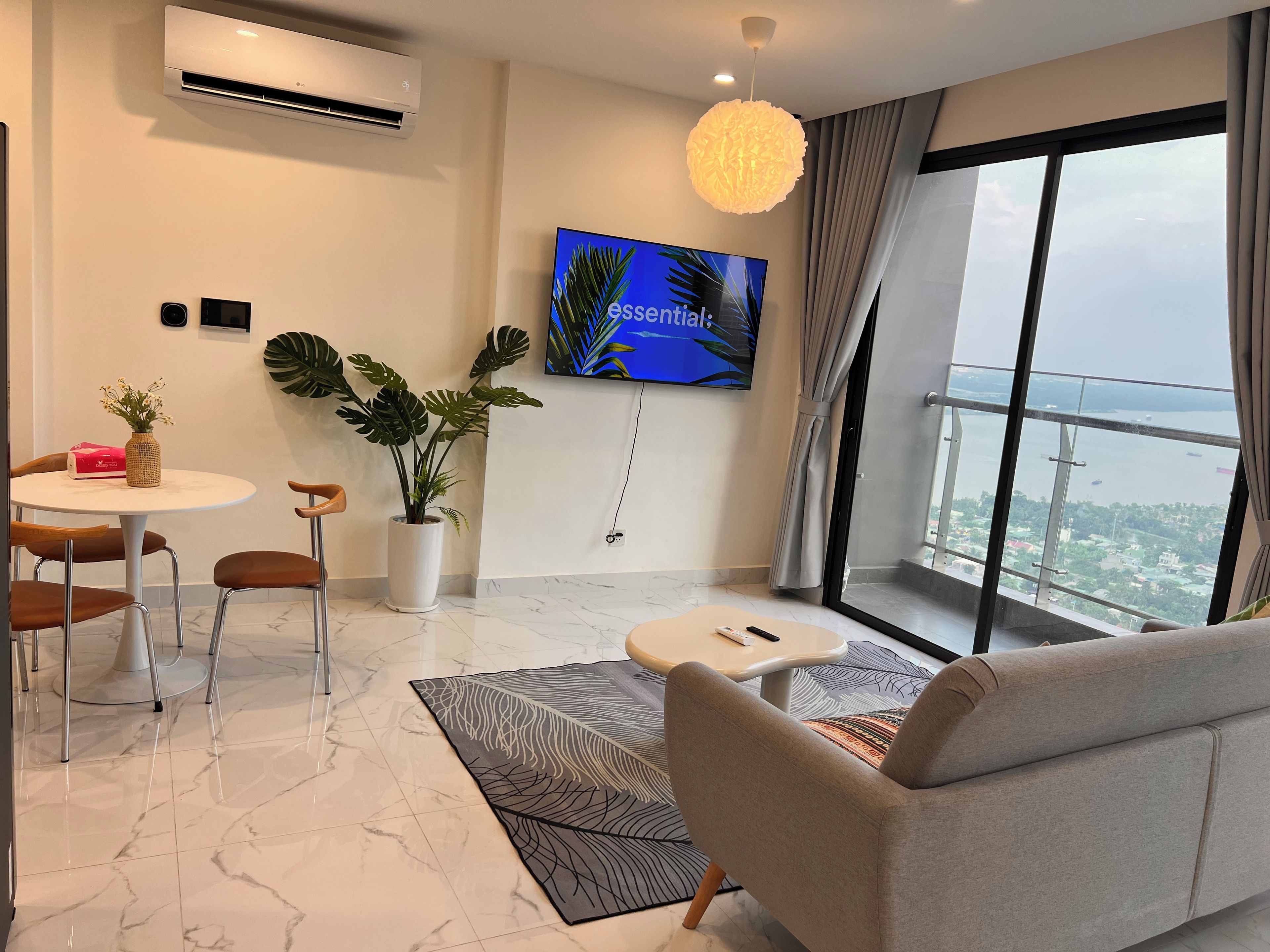 Uma sala de estar moderna com um sofá, uma mesa de jantar com cadeiras, uma planta em vaso, uma TV de tela plana e uma janela grande com vista.