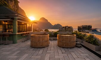 Terraço no telhado com barris de madeira, área de bar e vista para o mar e montanhas ao pôr do sol.