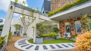 Um edifício moderno com fachada hexagonal única, um pátio central com caminho circular e diversos vasos de plantas.