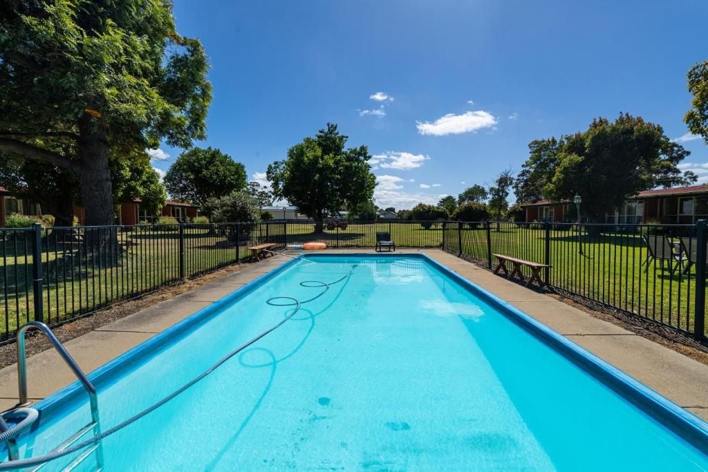 Piscina retangular com escada de metal, cercada por uma cerca preta, árvores e céu claro.