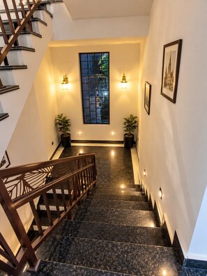 Escadaria com corrimão de madeira, piso de mármore preto e luminárias fixadas na parede.