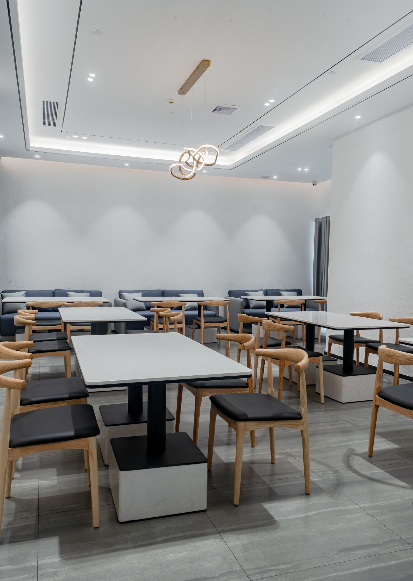 Uma área de refeições moderna, com várias mesas e cadeiras, um lustre no teto e uma abertura na parede.