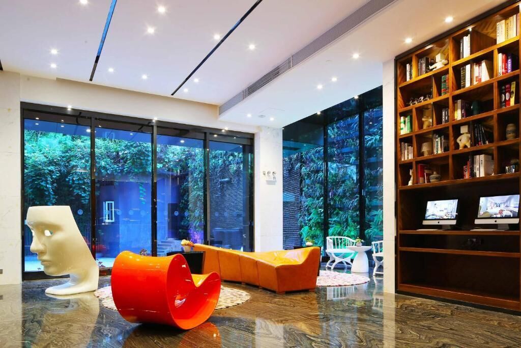Um lobby moderno com uma grande estante de livros, uma cadeira laranja única e um painel de vidro com vista para o verde.