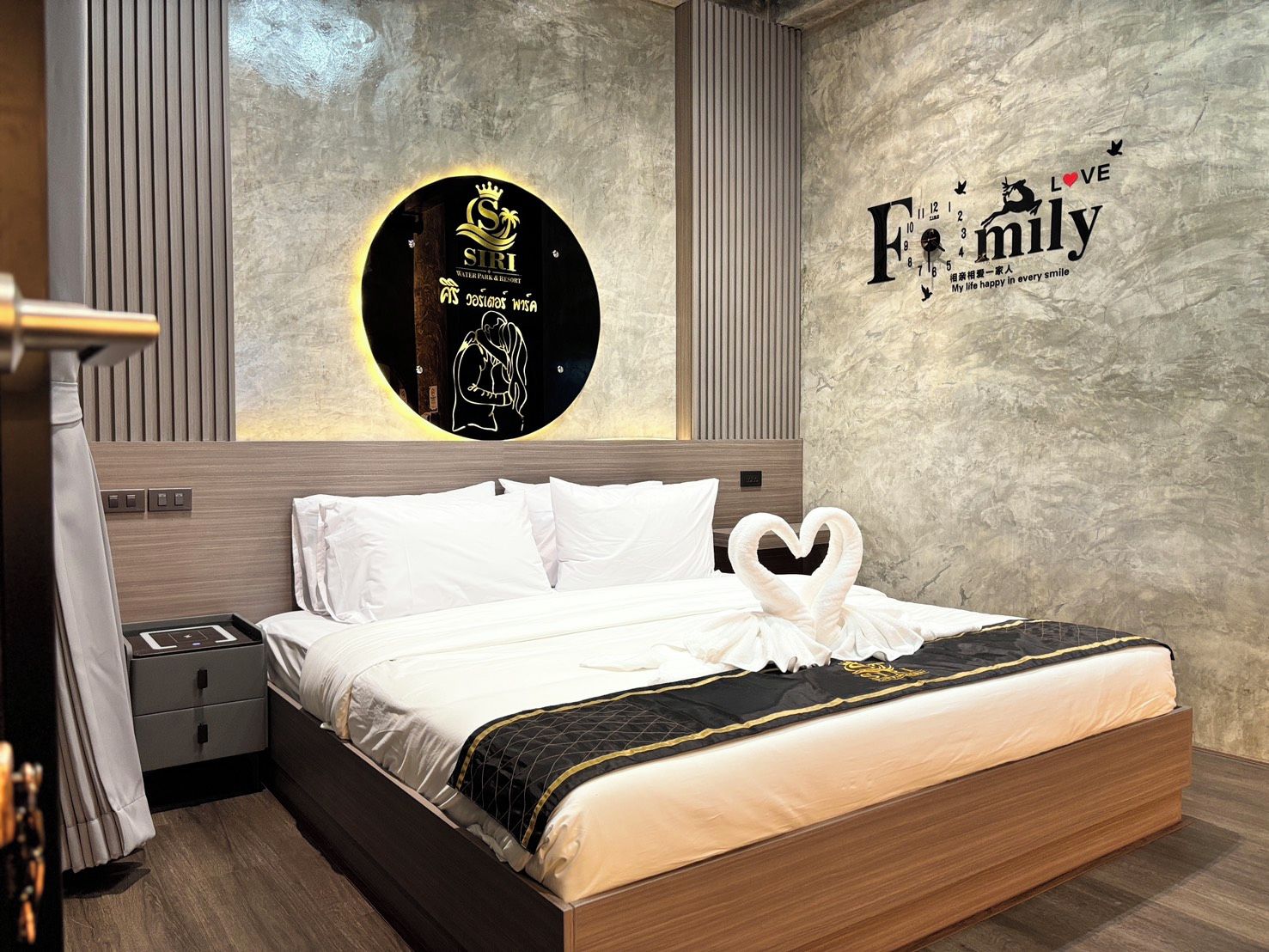 Quarto de hotel moderno com uma cama, uma mesinha de cabeceira, um relógio de parede e um logotipo circular na parede.