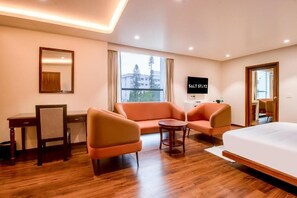 Quarto de hotel com cama, dois sofás, uma mesinha, escrivaninha, cadeira, televisão e janela com vista para edifícios e árvores.