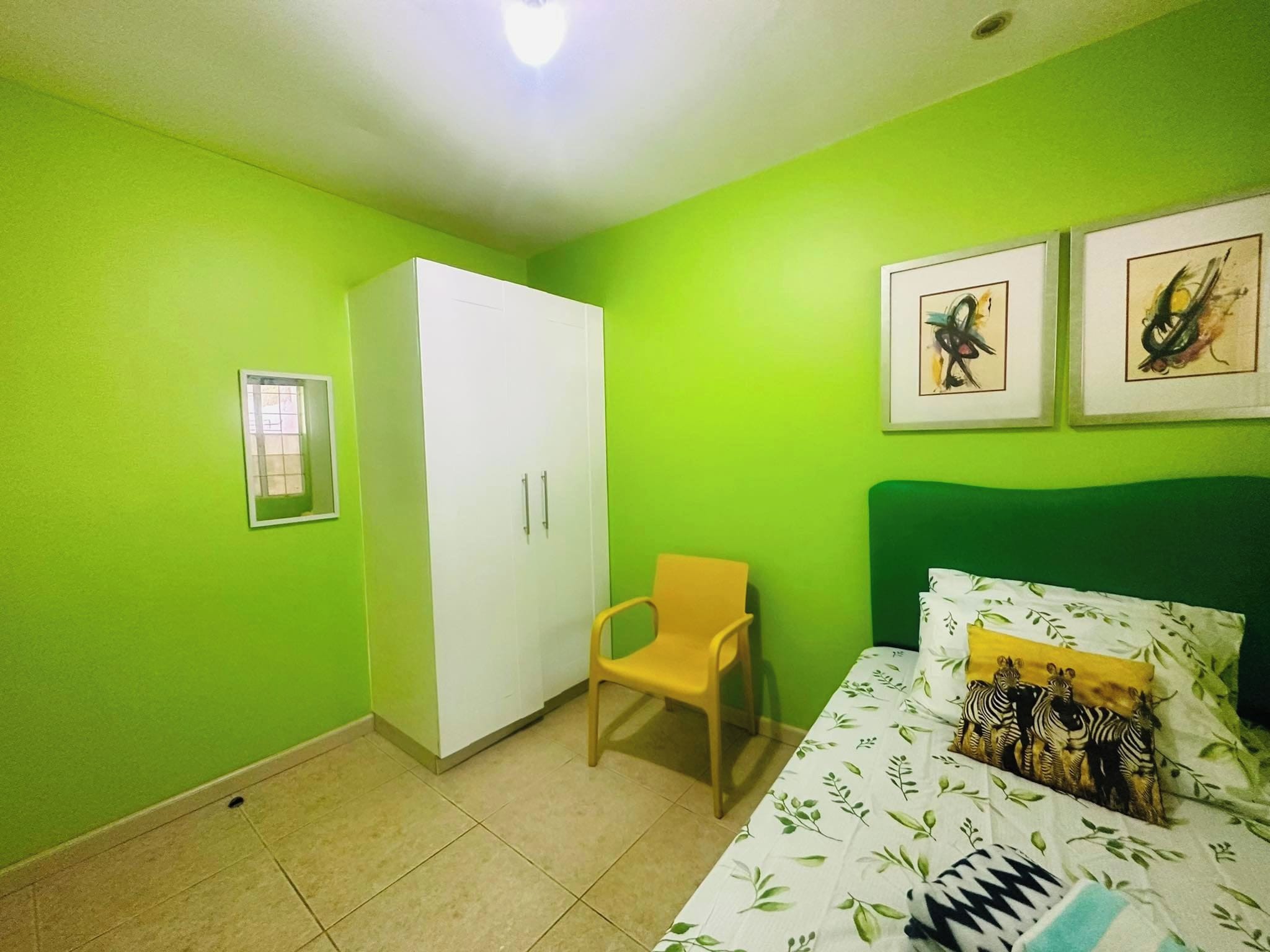 Quarto com paredes verdes, cama com roupa de cama estampada, guarda-roupa branco, janela pequena e duas obras de arte emolduradas.