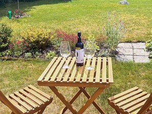 Mesa de madeira com duas cadeiras, uma garrafa de vinho e dois copos de vinho em um gramado com um jardim ao fundo.