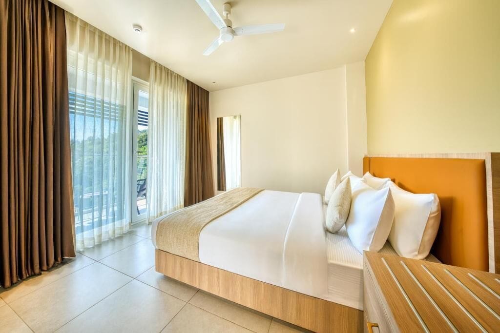 Quarto de hotel com cama, mesa de cabeceira de madeira, ventoinha no teto e vista para o verde lá fora.