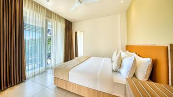 Quarto de hotel com cama, mesa de cabeceira de madeira, ventoinha no teto e vista para o verde lá fora.
