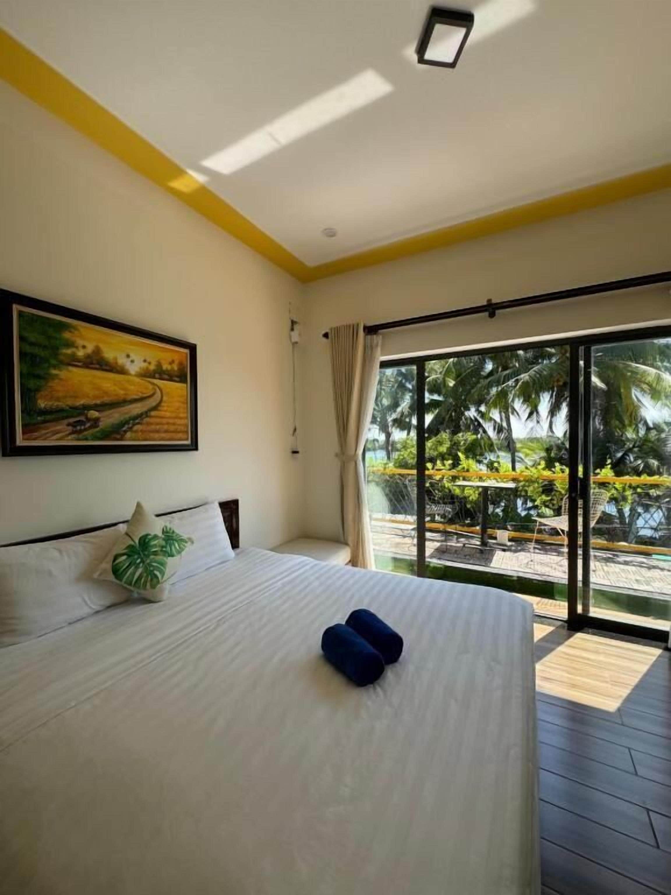 Quarto de hotel com uma cama grande, um quadro na parede e vista para palmeiras pela janela.
