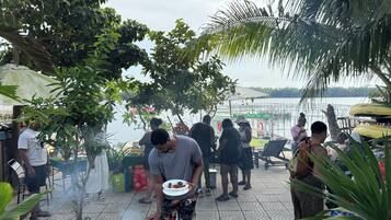 Área de refeições ao ar livre com churrasqueira, pessoas reunidas e vista para a água.