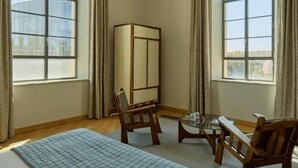 Quarto de hotel com uma cama grande, duas cadeiras, uma mesinha e vista da cidade por grandes janelas.