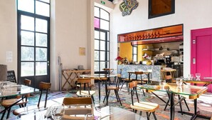 Uma sala de jantar ampla, com várias mesas e cadeiras, janelas grandes e uma cozinha visível por uma porta aberta.