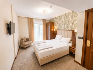 Quarto de hotel com cama, uma poltrona, televisão e parede com padrão de folhagens.